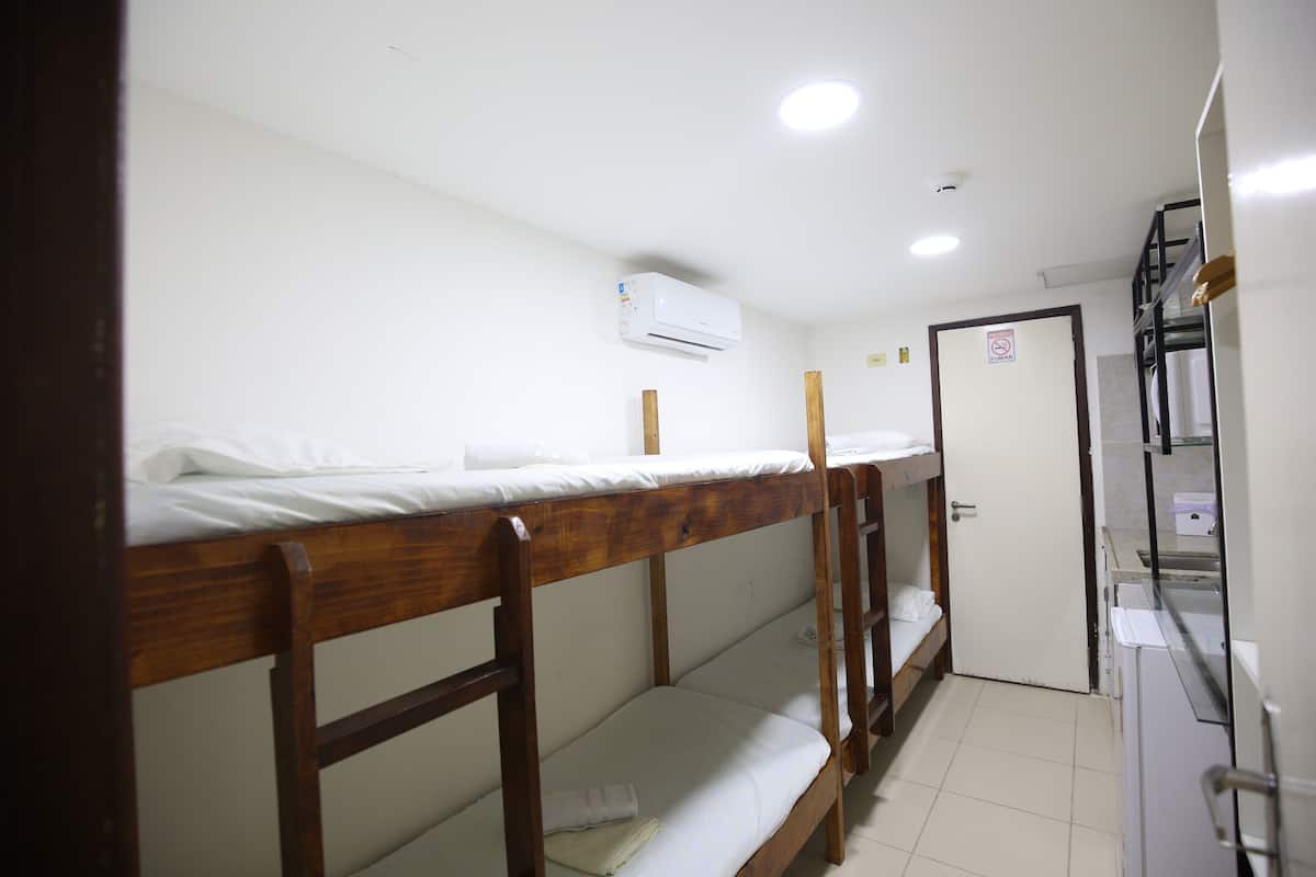 Quarto com beliches, ar condicionado branco, porta com placa de 'Não Fumar' e piso em azulejo.