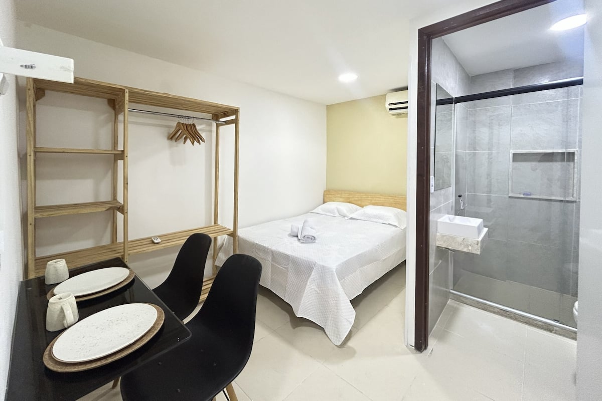 Quarto de hotel com cama, escrivaninha com cadeiras, espelho e chuveiro.
