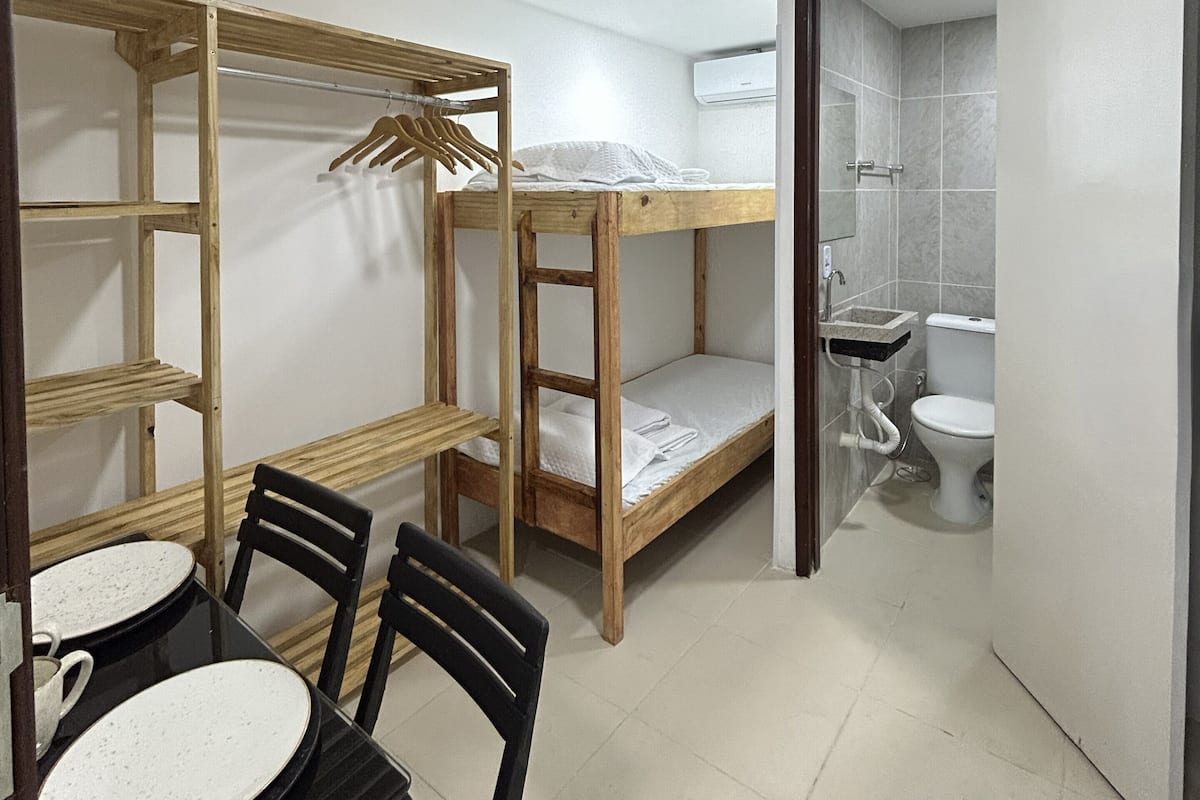 Quarto compacto com beliches, uma pequena mesa com cadeiras, pia e vaso sanitário.