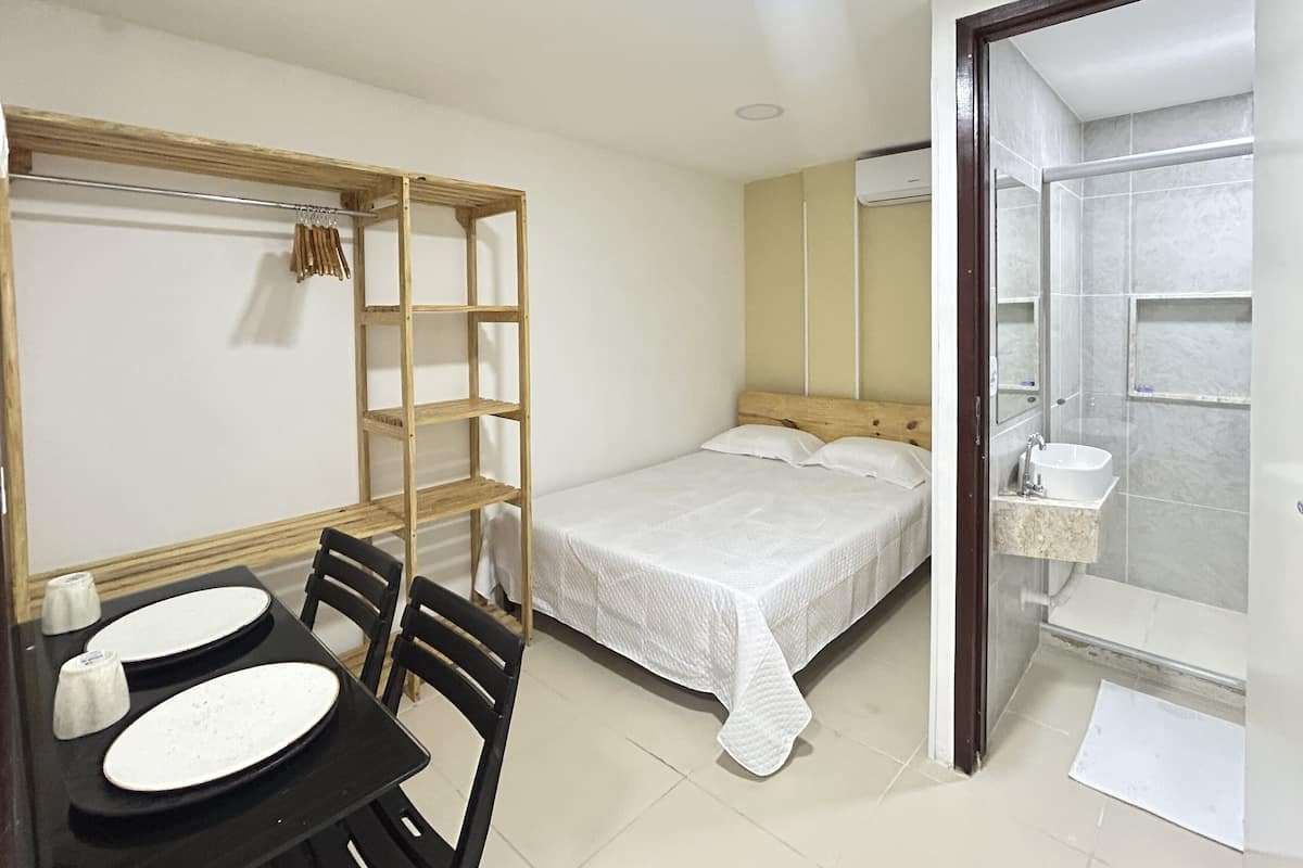 Quarto pequeno e limpo com uma cama, uma escrivaninha com cadeiras, uma prateleira de madeira e um banheiro com pia e chuveiro.