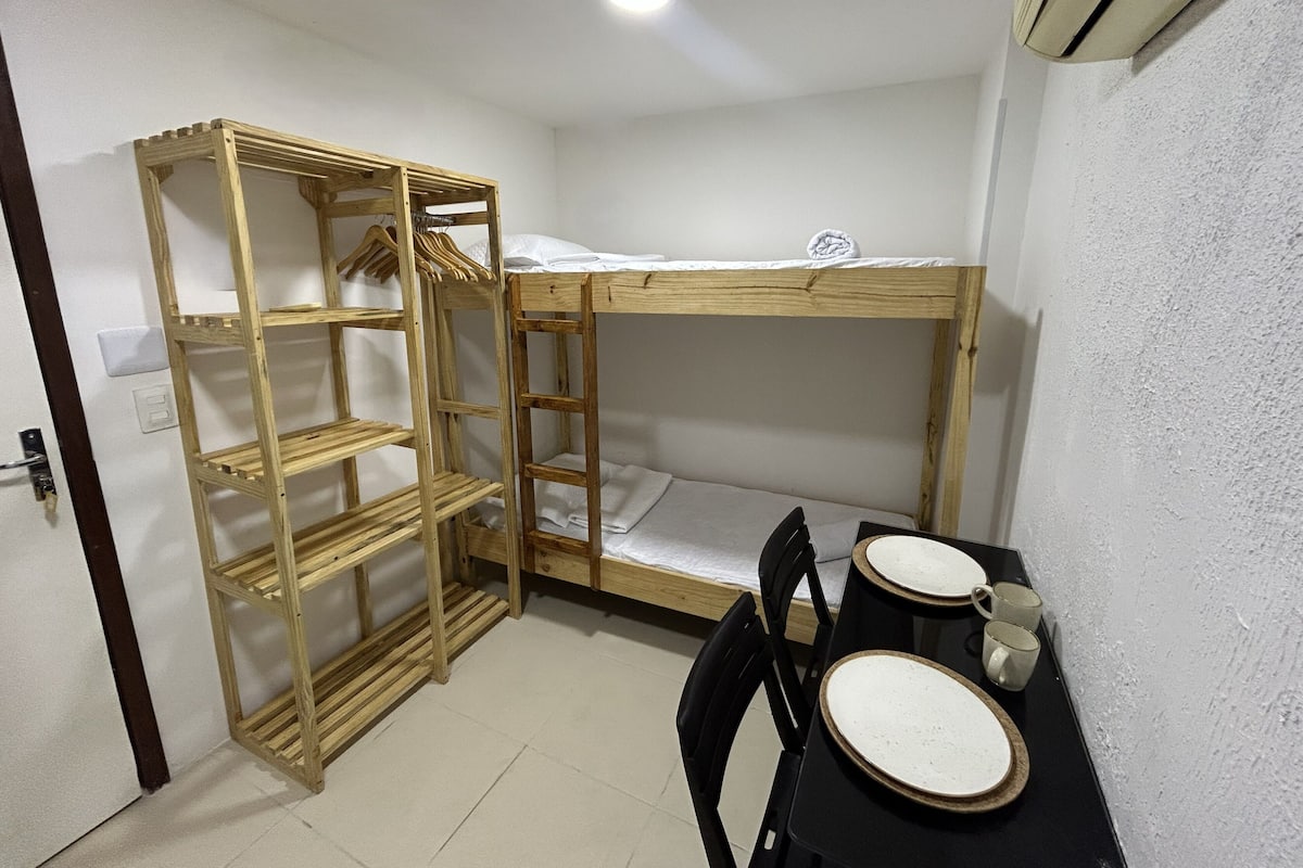 Quarto pequeno e compacto com beliche, estante de madeira, mesa com cadeiras e uma pequena mesa com xícaras.