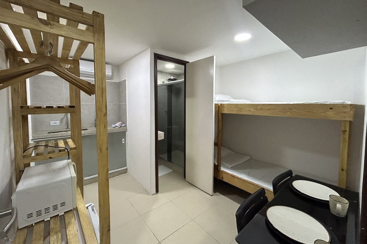 Quarto com beliches, prateleira de madeira, escrivaninha com cadeiras e banheiro com porta de vidro.