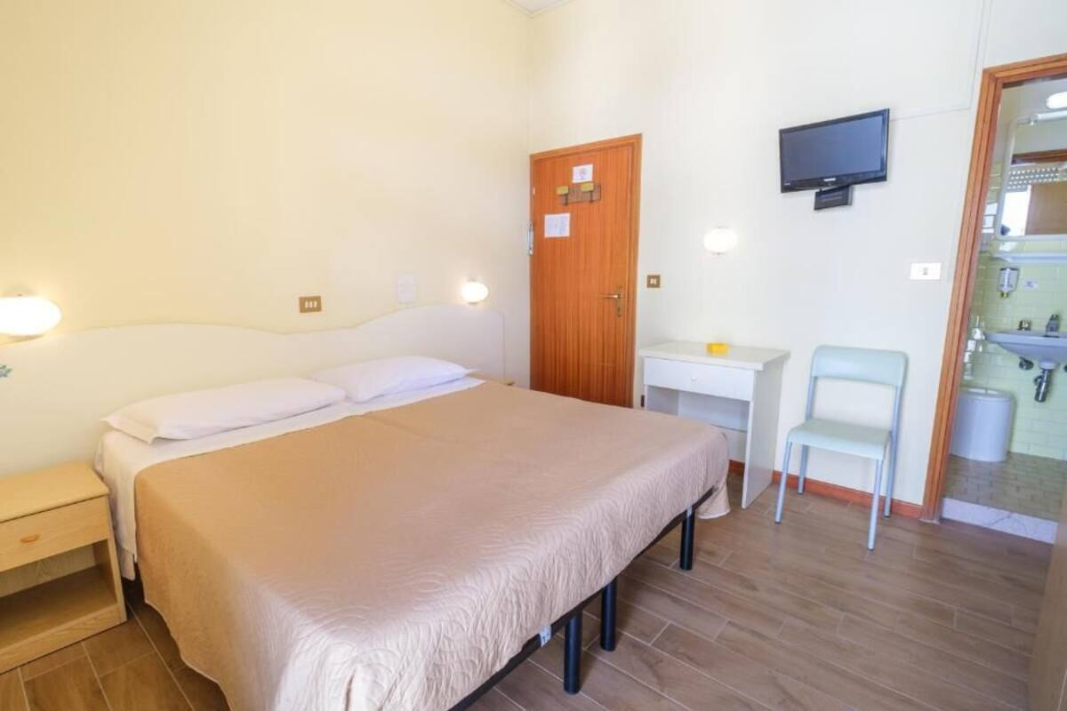 Quarto com cama, mesa de cabeceira, escrivaninha, cadeira e televisão na parede.