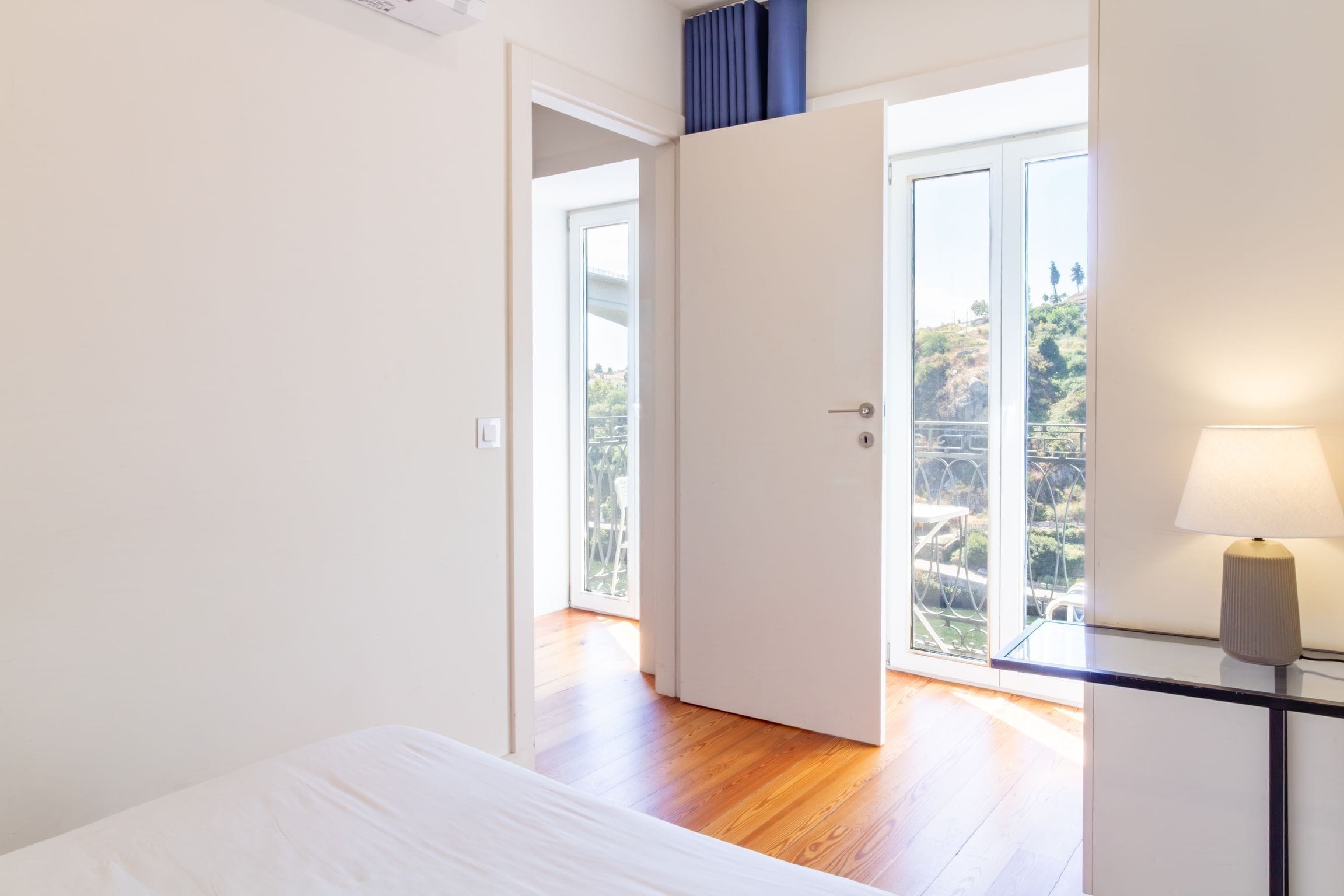 Quarto com cama, escrivaninha com luminária, porta de vidro deslizante para uma varanda e vista para o exterior.
