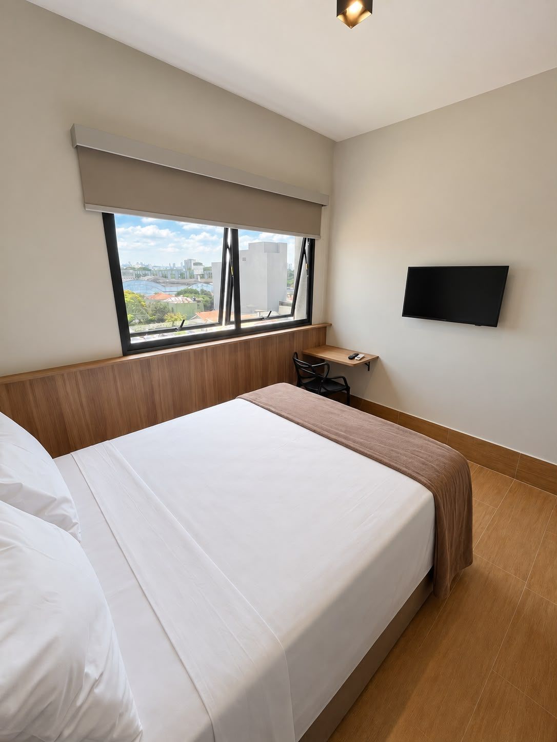 Quarto de hotel com uma cama, televisão na parede, janela com persianas e vista para a paisagem urbana.