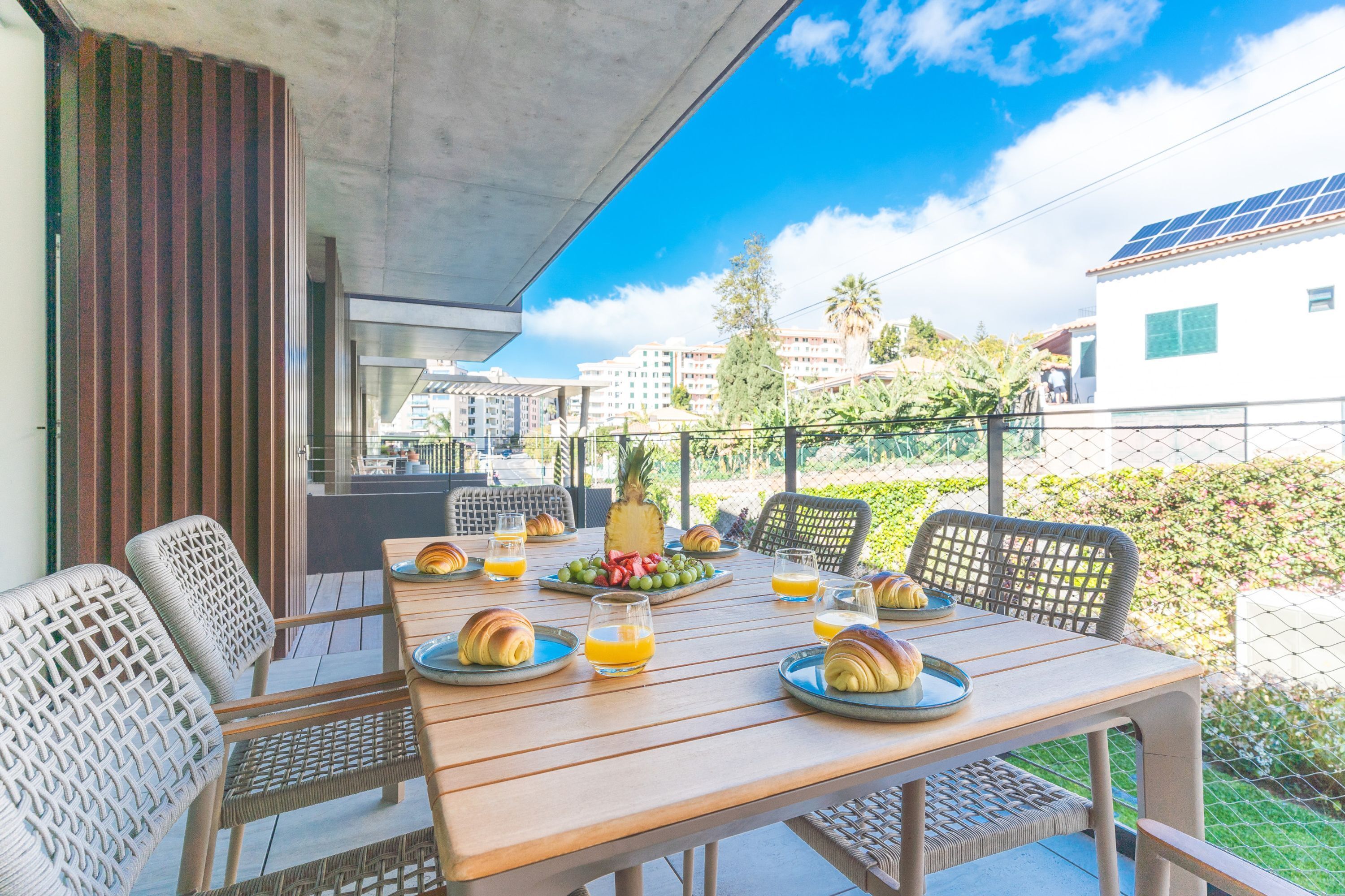 Uma área de refeições ao ar livre com mesa de madeira, cadeiras de vime e vista para edifícios residenciais.