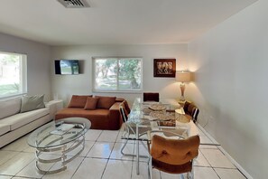 Uma sala de estar moderna com mesa de centro de vidro, sofá marrom e uma área de jantar com mesa e cadeiras de vidro.