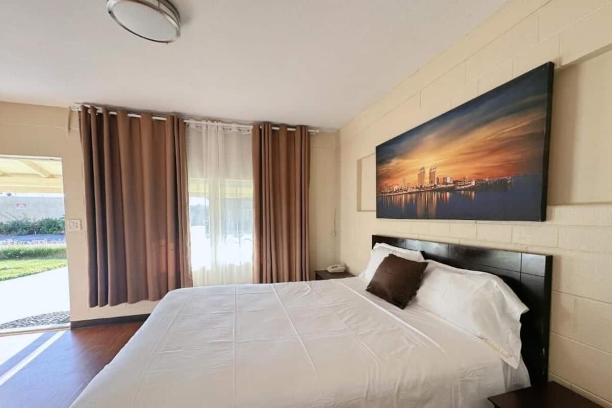 Quarto de hotel com cama grande, mesa de cabeceira, pintura de uma paisagem urbana e vista para o exterior.
