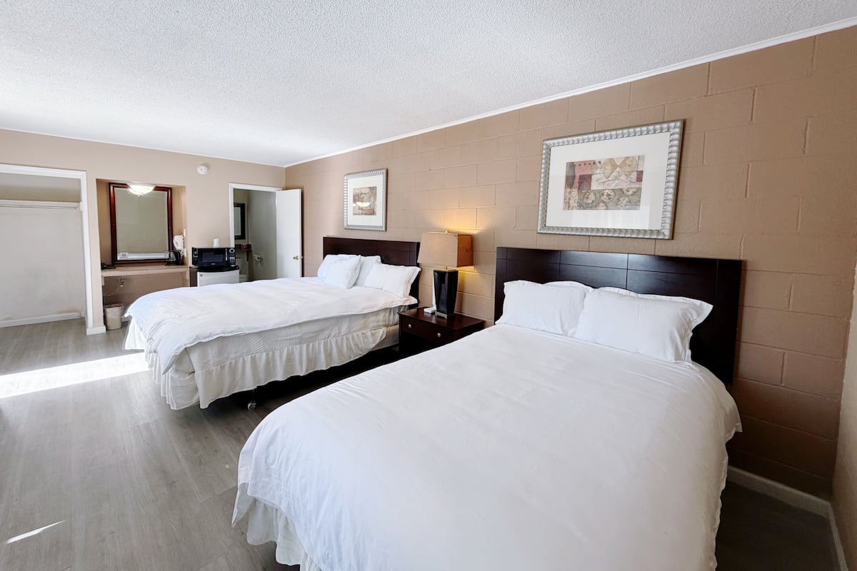 Quarto de hotel com duas camas, uma escrivaninha, uma cadeira, um espelho e um quadro na parede.