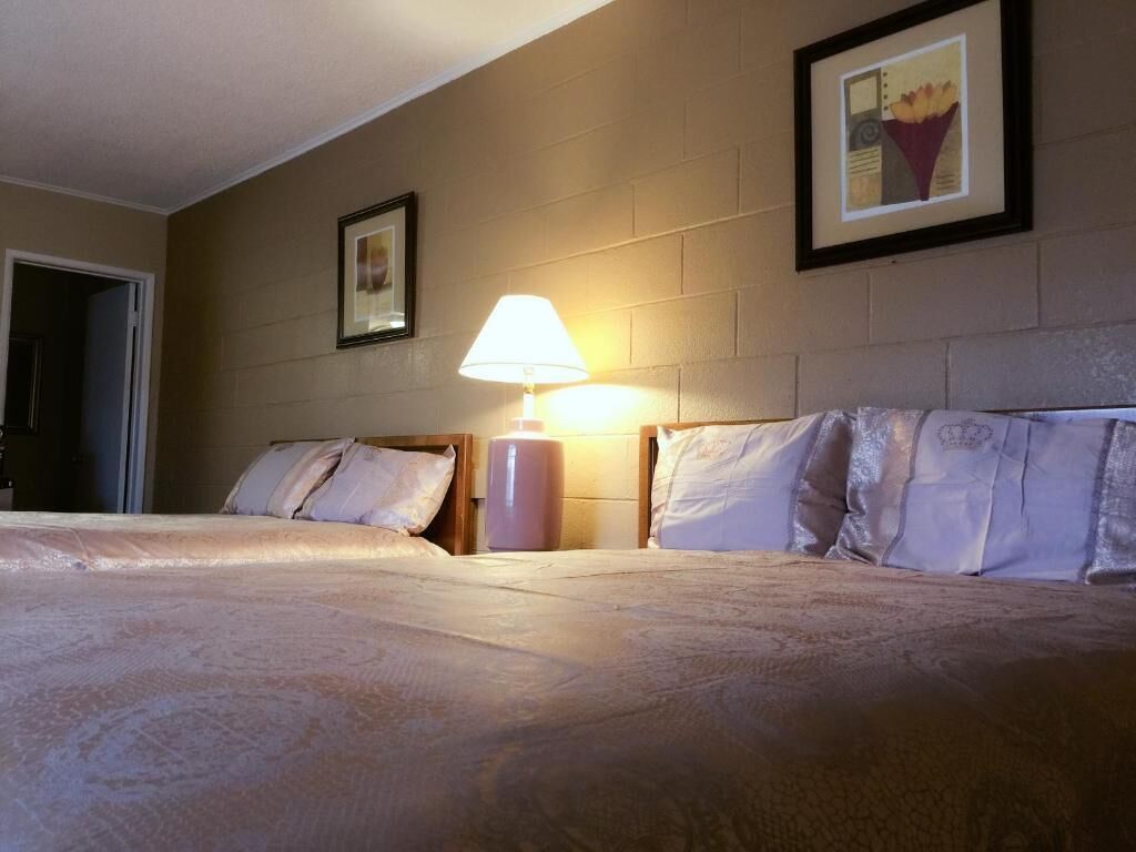 Quarto de hotel com duas camas, abajur, quadro na parede e uma porta à esquerda.