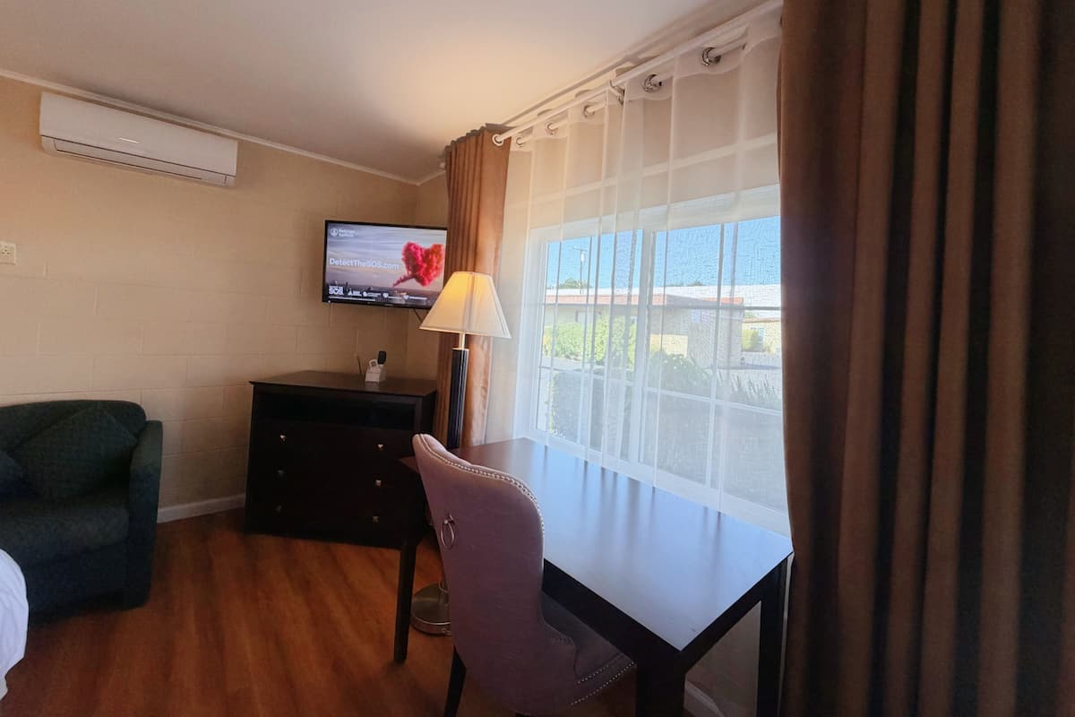 Quarto de hotel com televisão, escrivaninha com luminária, cadeira, sofá e janela com cortinas.