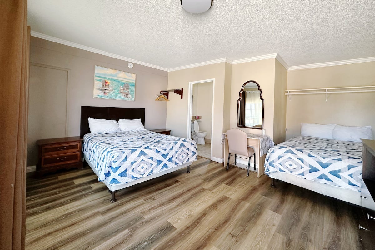 Quarto de hotel com duas camas, uma escrivaninha, uma cadeira, um espelho e um quadro na parede.