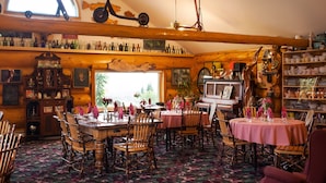 Um interior rústico com vigas de madeira, área de jantar com mesas e cadeiras redondas, e diversos objetos decorativos nas prateleiras.