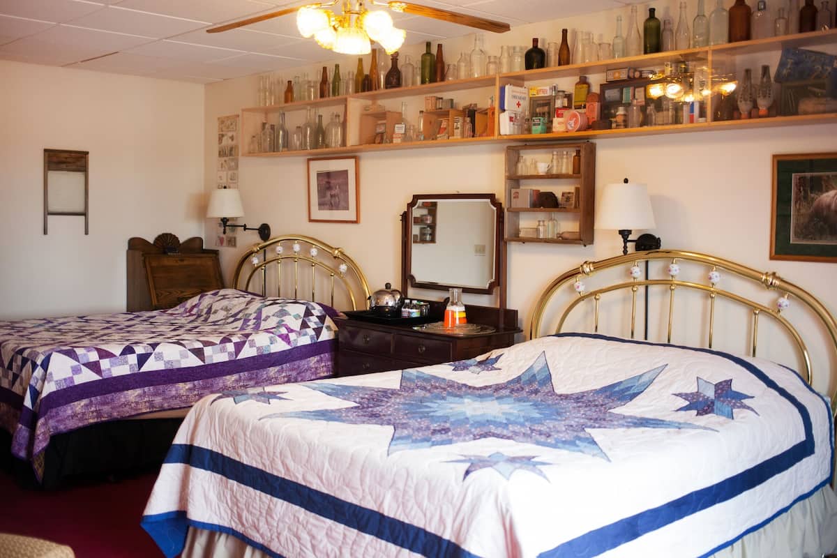 Quarto com duas camas, cabeceira de madeira, prateleira com garrafas e um espelho.