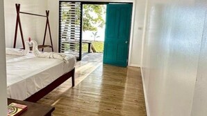 Quarto com cama, piso de madeira, porta verde e vista pela janela.
