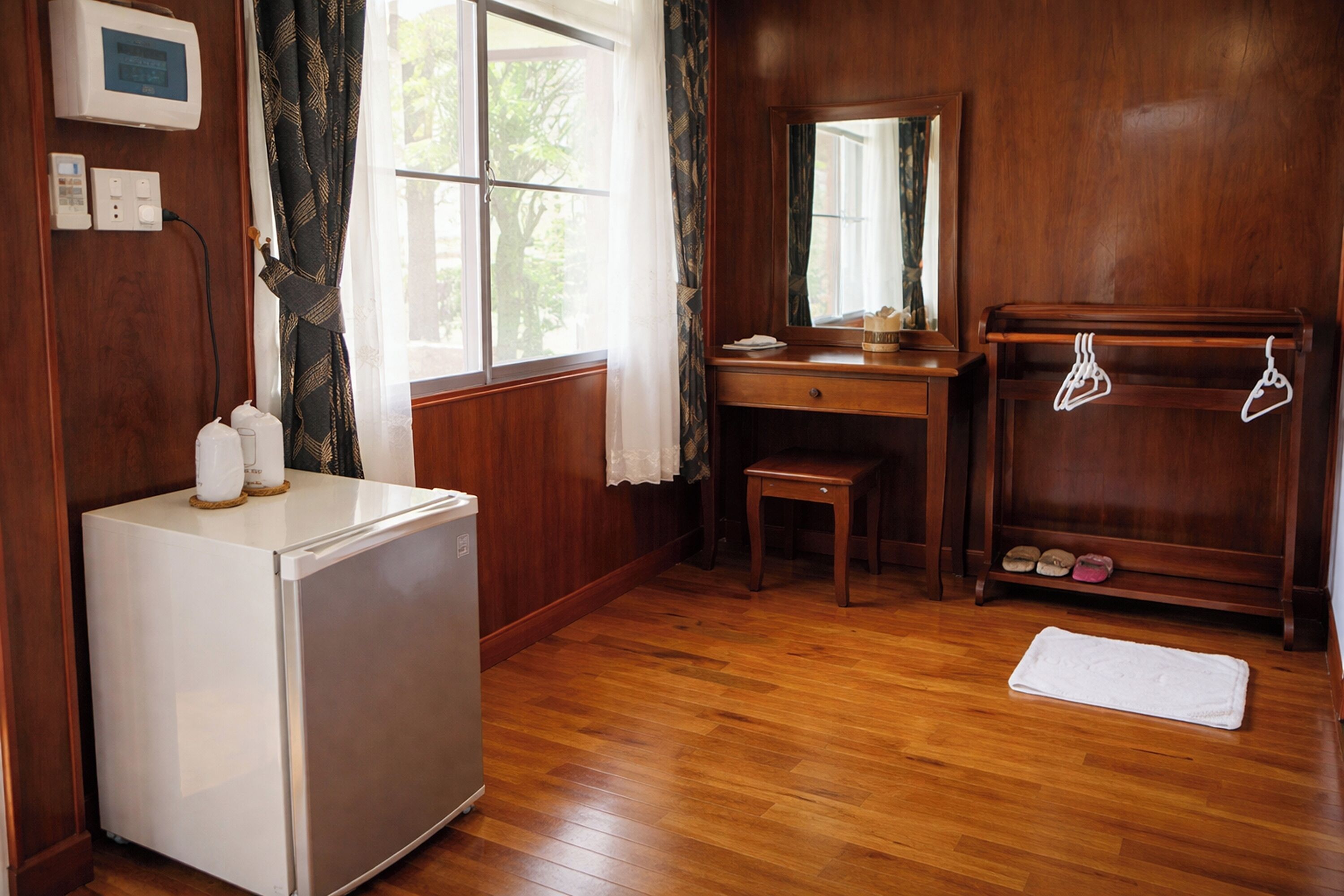 Quarto com piso de madeira, uma pequena geladeira branca, escrivaninha de madeira, espelho e sapateira.