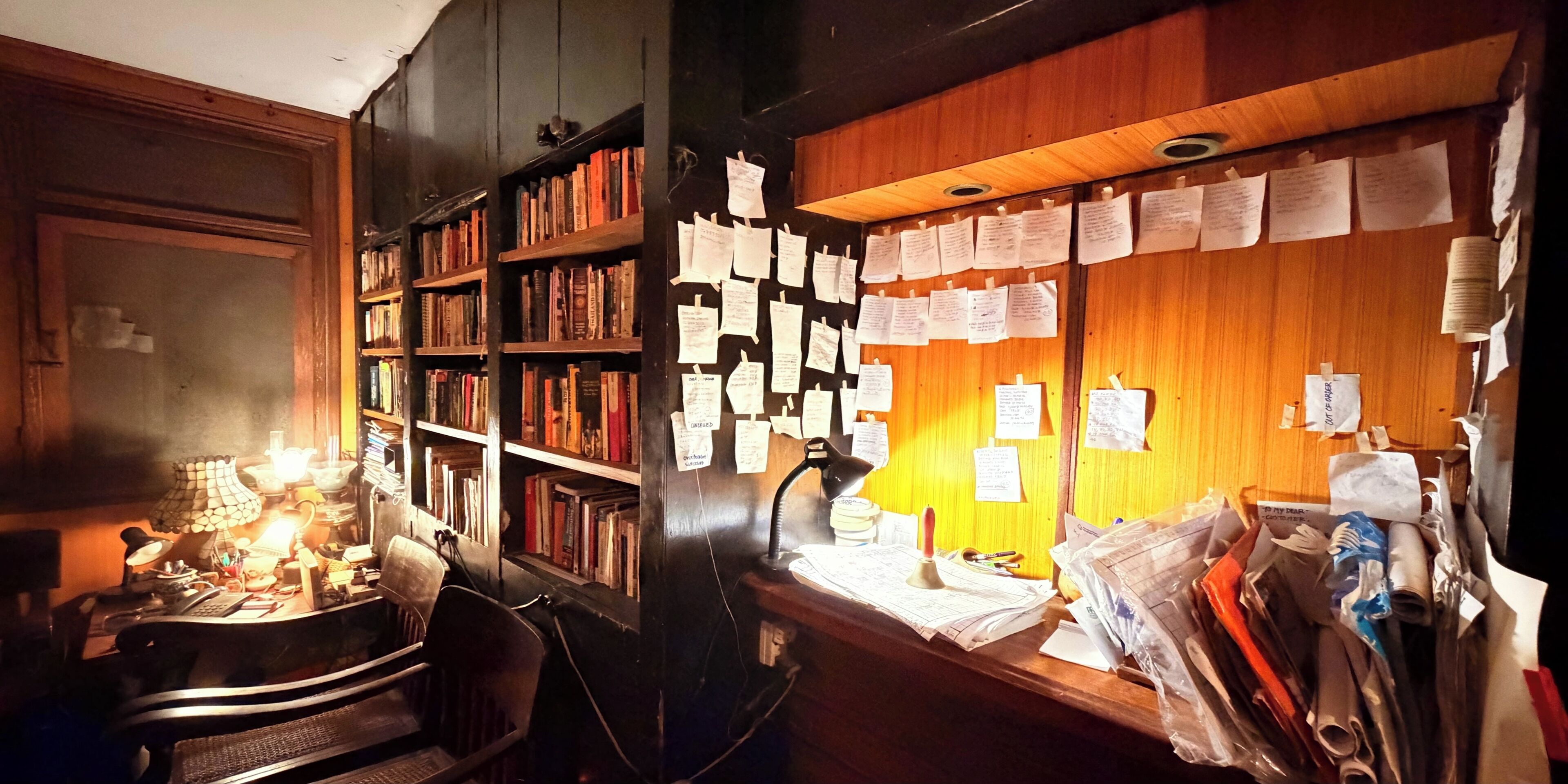 Quarto desarrumado com escrivaninha, cadeira, luminária e estantes repletas de livros. Papéis estão fixados na parede.