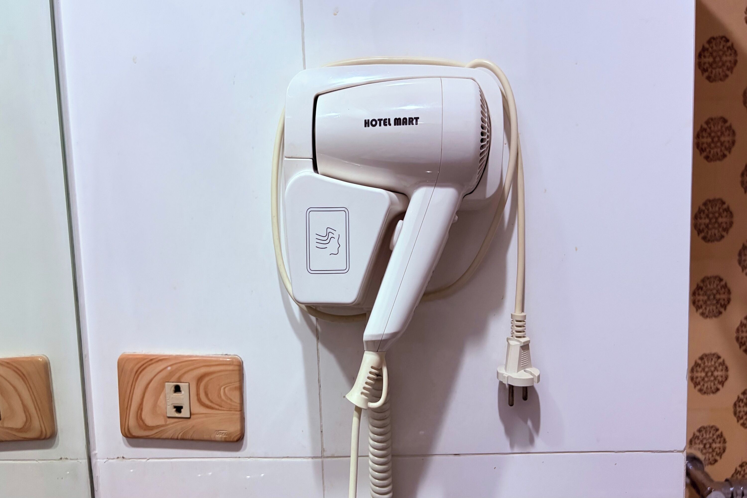 Secador de cabelo branco com fio conectado a uma tomada, identificada como 'HOTEL MART'.