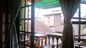 Um quarto com uma mesa e cadeiras de madeira, janela com vista para prédios, e um toldo verde.