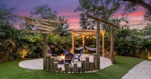 Um gazebo de madeira com lareira e cadeiras, rodeado por um jardim e um poste de sinalização indicando direções para vários locais.