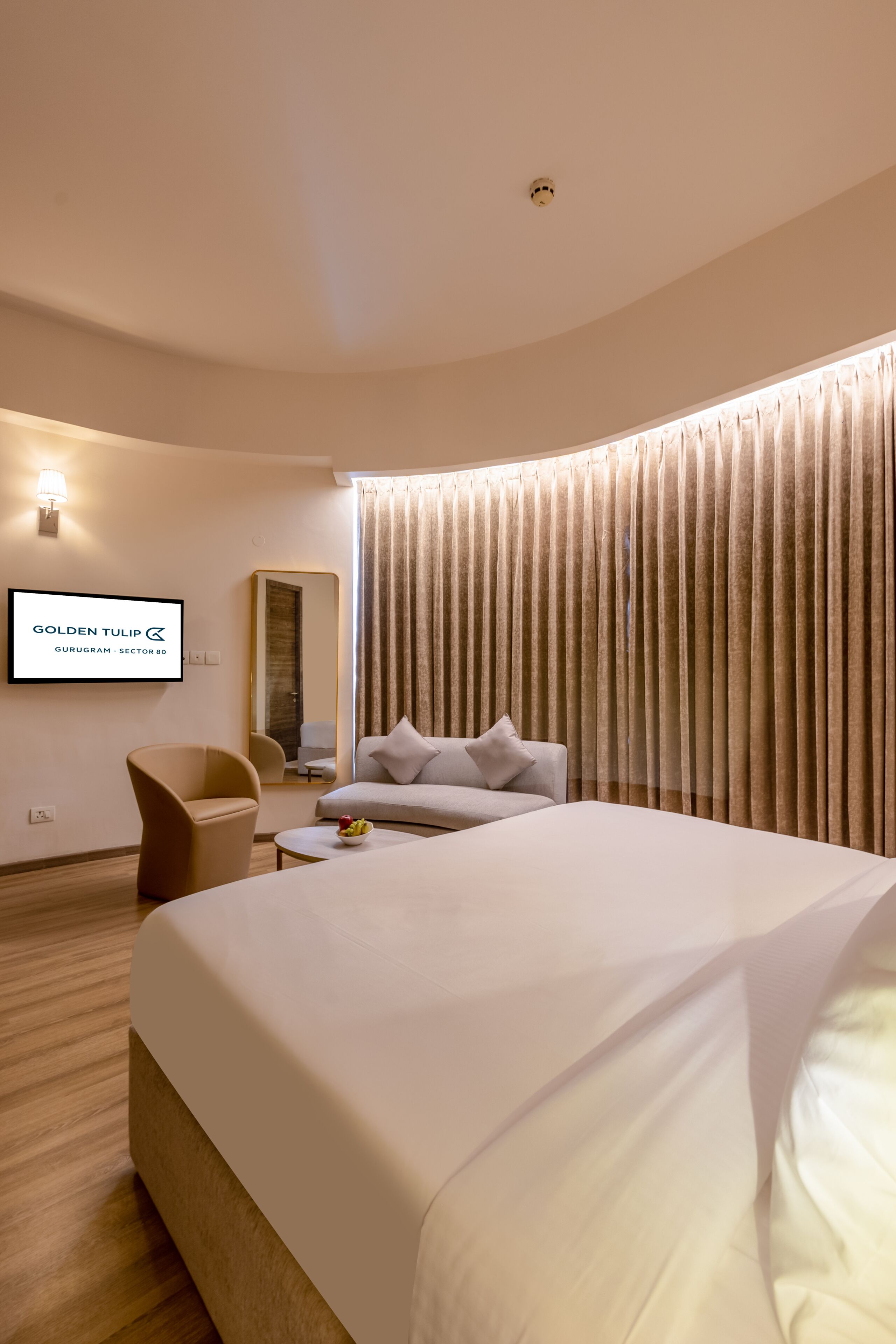 Quarto de hotel com uma cama grande, uma cadeira, uma pequena mesa com uma tigela de frutas, um espelho e uma TV na parede exibindo 'GOLDEN TULIP GURUGRAM - SECTOR 80'.