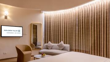Quarto de hotel com uma cama grande, uma cadeira, uma pequena mesa com uma tigela de frutas, um espelho e uma TV na parede exibindo 'GOLDEN TULIP GURUGRAM - SECTOR 80'.