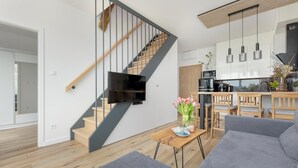 Uma sala de estar moderna com escada, sofás cinzas, mesa de centro de madeira e área de cozinha com armários de madeira e luminárias pendentes.
