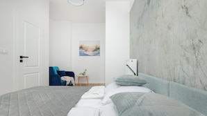 Quarto com uma cama, uma cadeira, uma mesinha com uma planta e um quadro na parede.