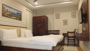 Quarto com cama, guarda-roupa de madeira, escrivaninha, cadeira e televisão fixada na parede.