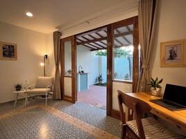 Quarto com escrivaninha de madeira, uma cadeira, um laptop, uma mesinha com uma planta e vista para uma cozinha e área externa.
