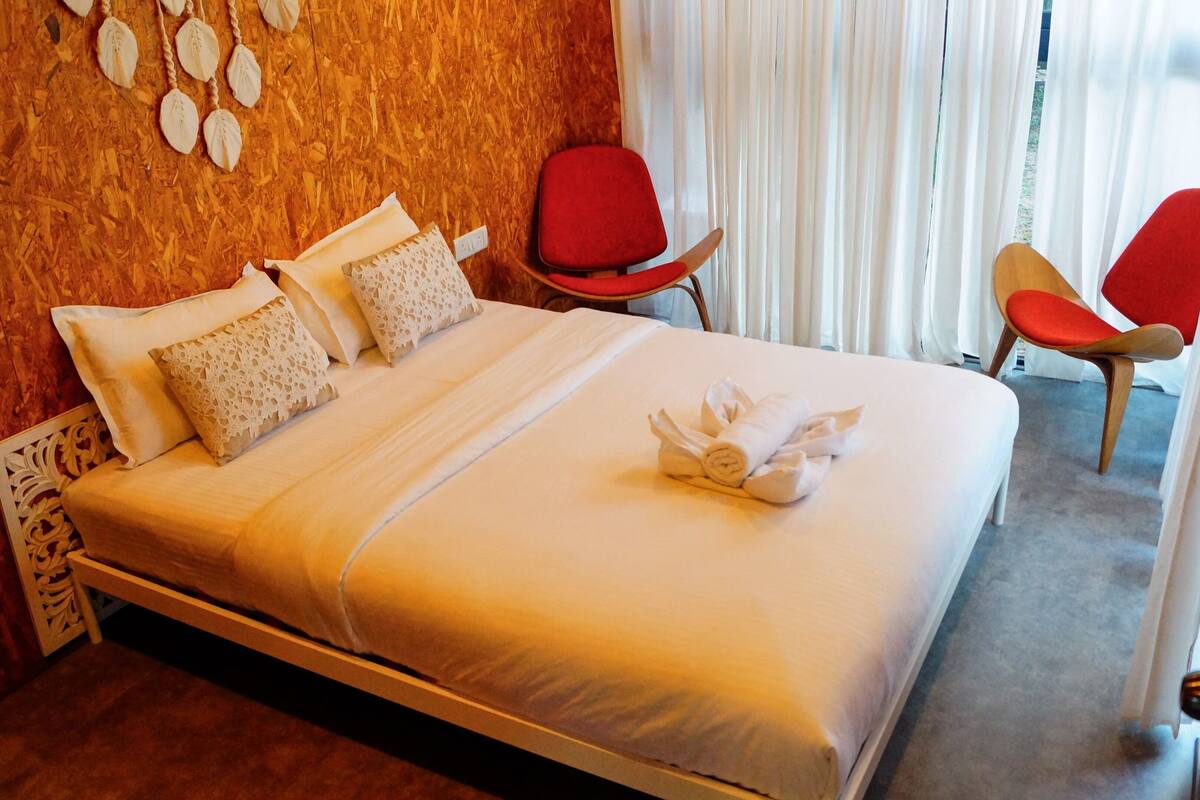 Quarto de hotel com uma cama, duas cadeiras vermelhas, uma parede de madeira e uma cortina branca.