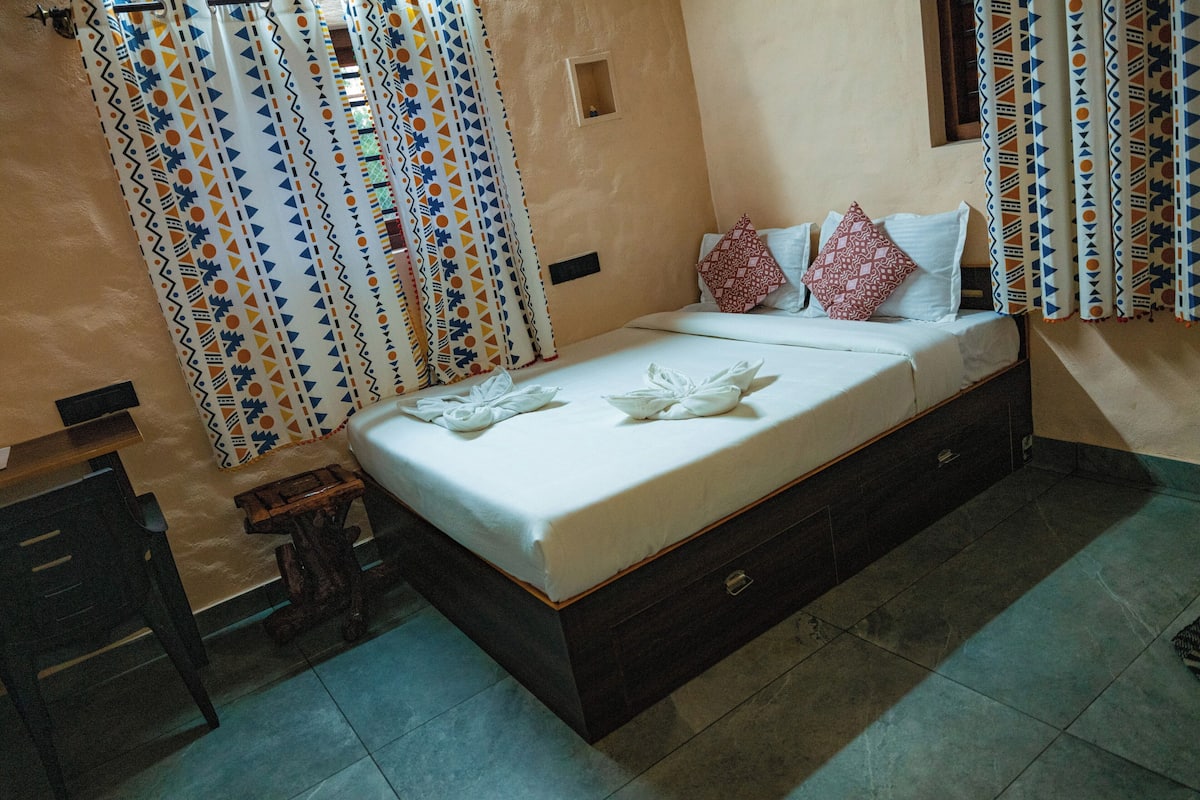Cama bem arrumada com almofadas, mesa de cabeceira de madeira, uma pequena mesa e uma janela com cortinas.