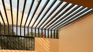 Quarto com teto inclinado, área de estar com almofadas, um galo decorativo e vista por uma janela com grades.