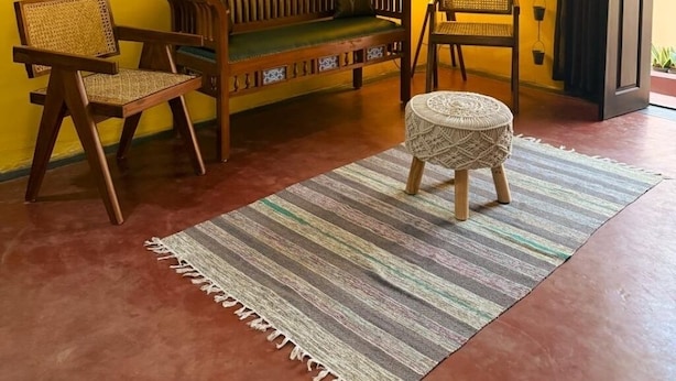 Um quarto com parede amarela, sofá de madeira, duas cadeiras de vime, um banquinho trançado e um tapete listrado.