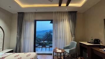 Quarto de hotel com cama, escrivaninha, cadeira, mesa pequena com um copo e varanda com vista para as colinas.