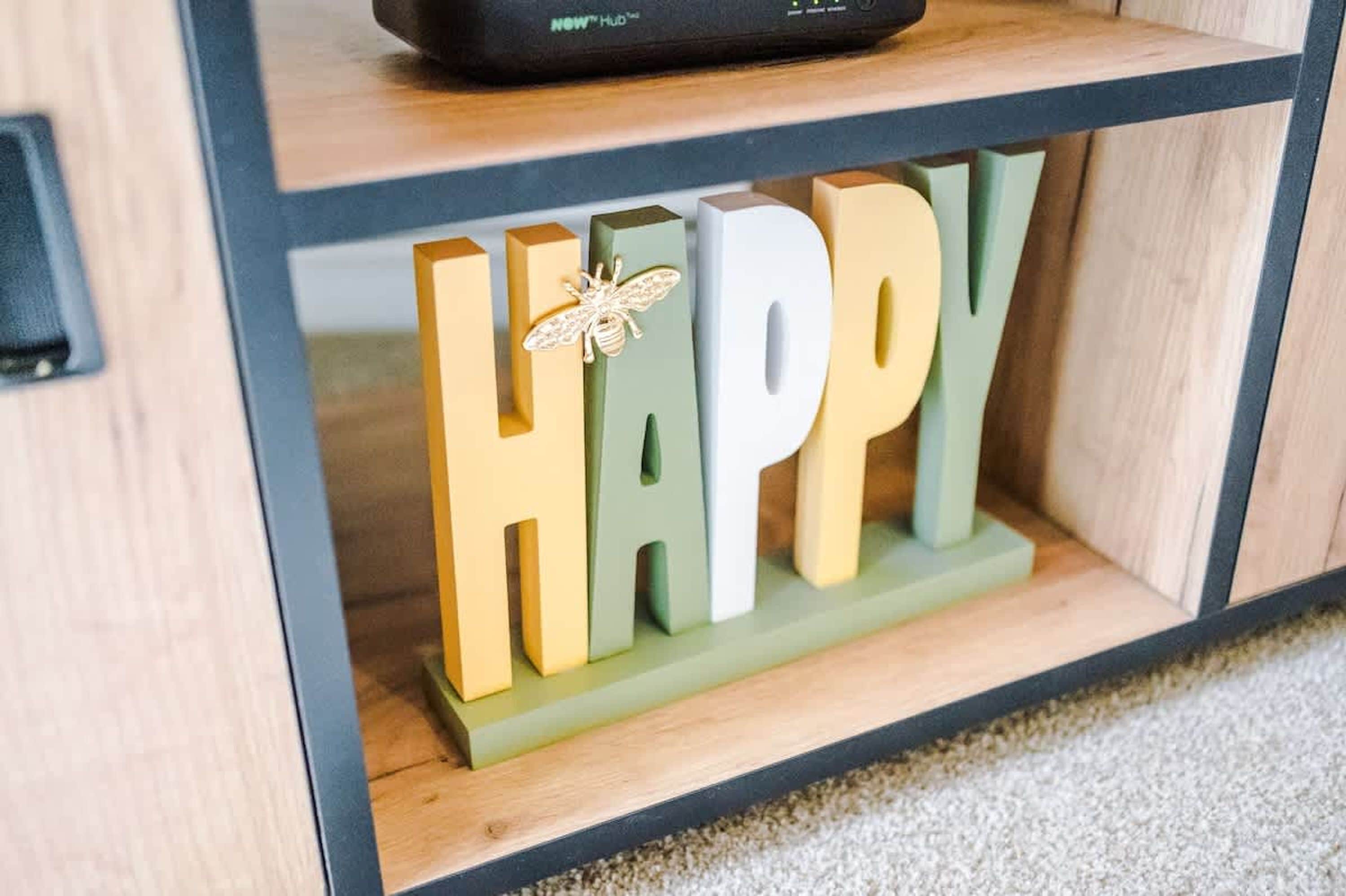 Uma prateleira de madeira com um aparelho eletrônico preto em cima e a palavra 'HAPPY' em letras coloridas com um enfeite de abelha.