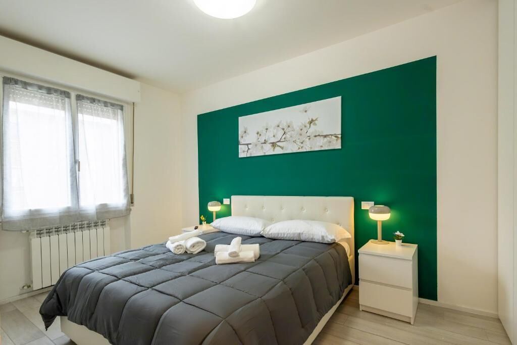 Quarto com cama, mesa de cabeceira, luminária na parede, radiador e janela com cortinas.