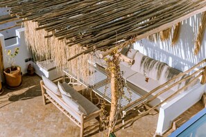 Uma área externa iluminada pelo sol com assentos, um pergolado, cadeiras de descanso e uma parede branca.