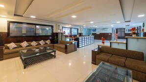 Um hall de hotel com balcão de recepção, sofás, mesa de centro e uma tela de televisão.