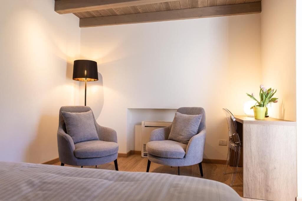 Quarto com duas poltronas cinzentas, uma escrivaninha com planta em vaso, luminária e cama.