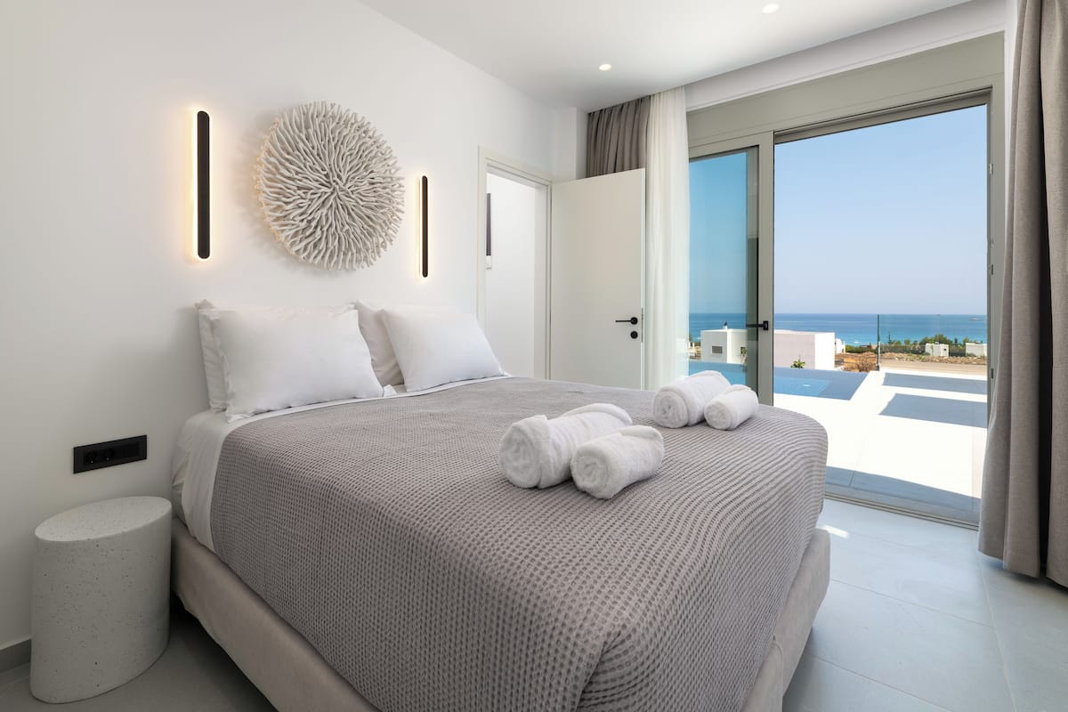 Quarto moderno com uma cama grande, uma decoração circular na parede e vista para o mar através de uma porta aberta.