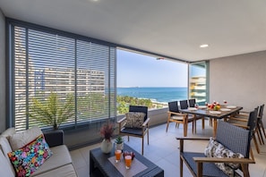 Uma varanda com vista para o mar, equipada com mesa, cadeiras e um sofá com almofada colorida.