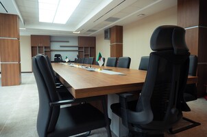 Uma sala de conferências com uma mesa comprida, cadeiras pretas, uma bandeira e uma televisão na parede.