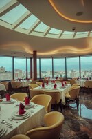 Restaurante com mesas redondas, cadeiras amarelas e vista para a cidade através de grandes janelas.