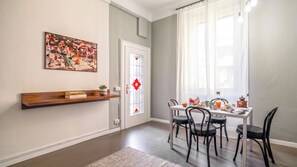 Uma sala de jantar com uma mesa branca, cadeiras pretas, uma estante de madeira com livros e um quadro na parede.