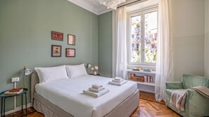 Quarto com cama, luminárias de cabeceira, janela com cortinas, mesa de cabeceira com um livro e uma poltrona verde.