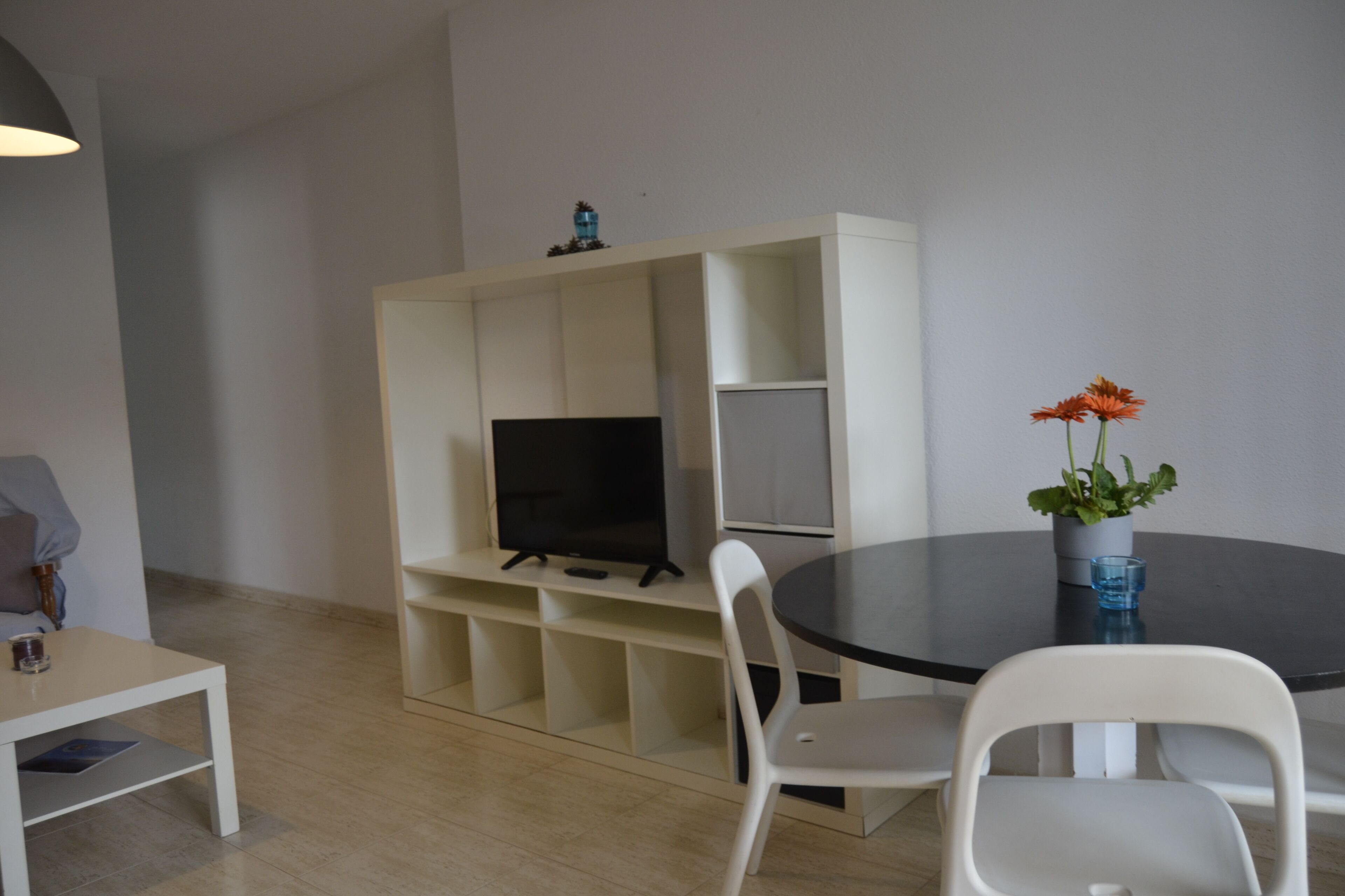 Quarto moderno com televisão de tela plana, cadeiras brancas, mesa redonda e planta em vaso.