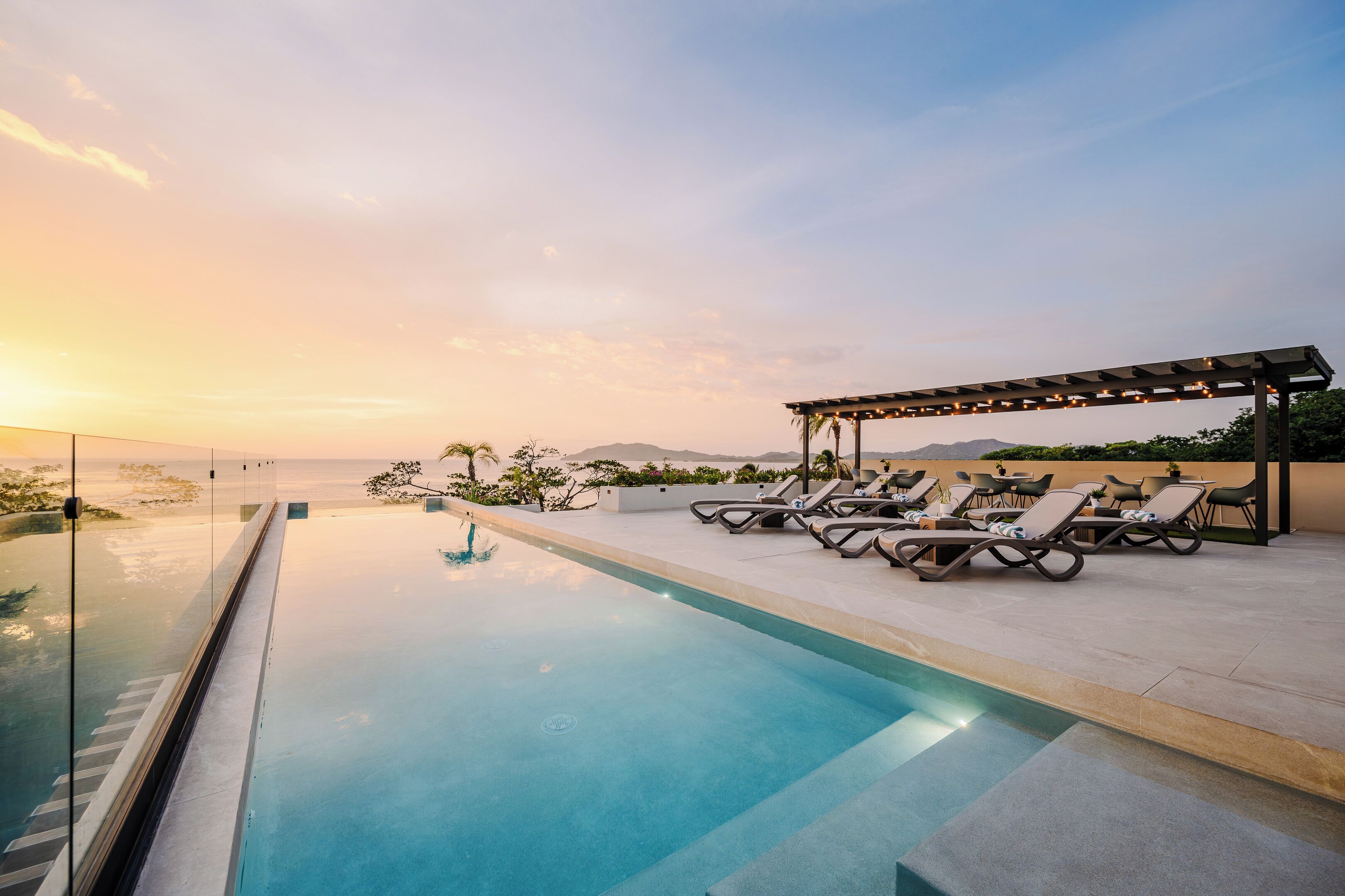 Piscina infinita no terraço com espreguiçadeiras e pérgola, com vista para um corpo de água e montanhas ao longe.