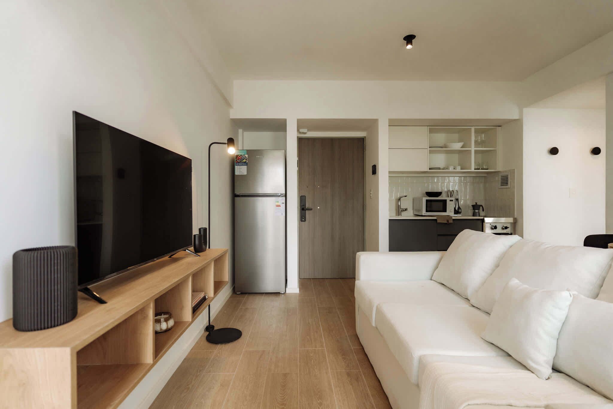 Uma sala de estar moderna com televisão de tela plana, móvel de entretenimento de madeira, sofá branco e uma área de cozinha ao fundo.