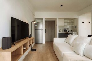 Uma sala de estar moderna com televisão de tela plana, móvel de entretenimento de madeira, sofá branco e uma área de cozinha ao fundo.